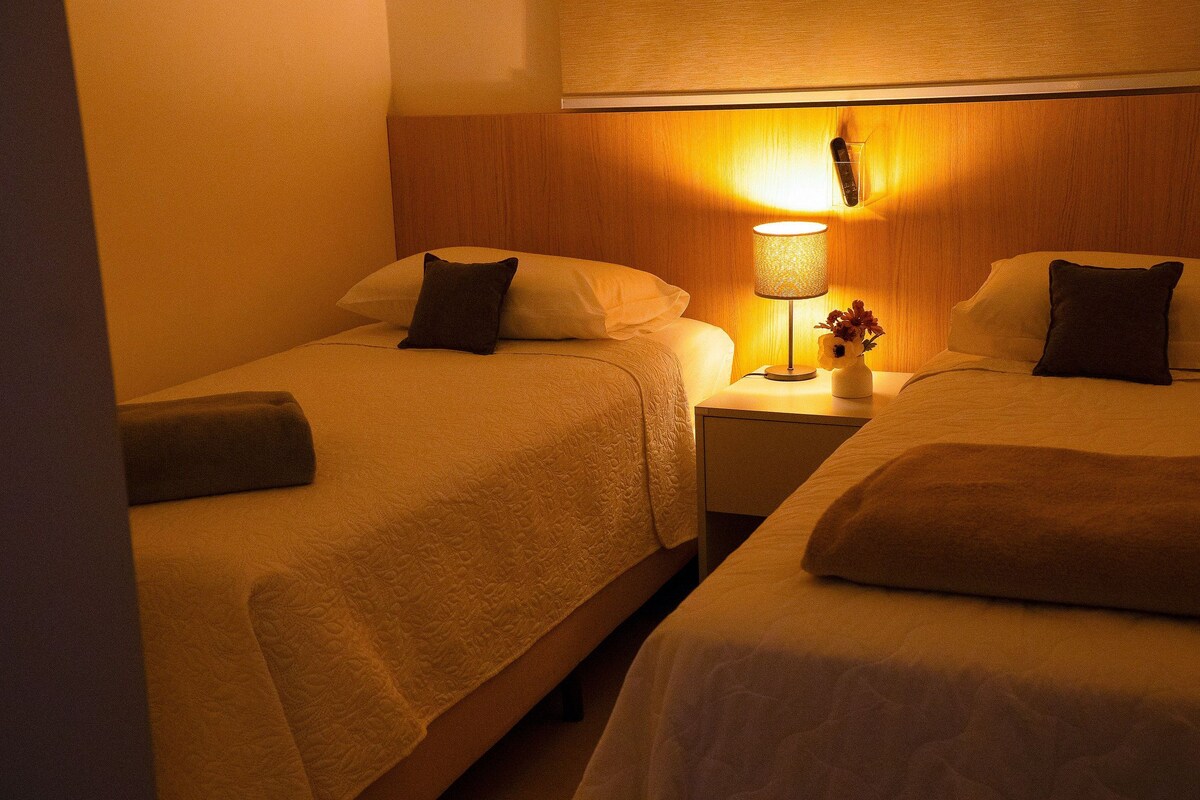 Quarto de hotel com duas camas, uma mesa de cabeceira com abajur e um vaso com flores.