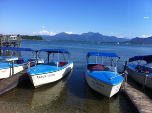 Uma fileira de barcos pequenos atracados em um cais, com água cristalina e montanhas ao fundo.