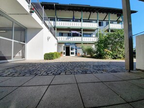 Entrada de hotel com um pátio pavimentado, uma varanda com grades e uma placa com a inscrição 'Appart-Hotel'.