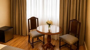 Um quarto com uma mesa redonda de madeira, duas cadeiras, um pequeno armário escuro e uma janela com cortinas bege.