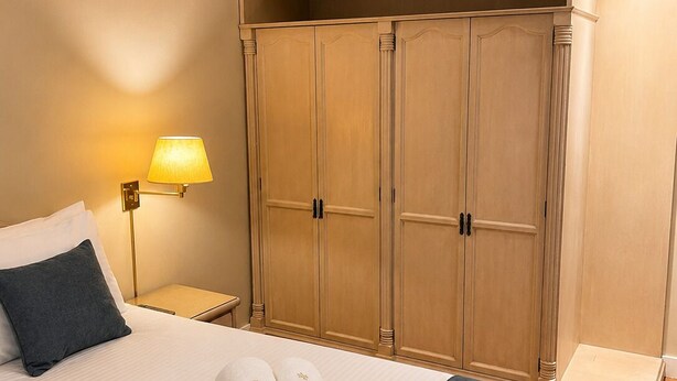 Quarto de hotel com uma cama grande, um guarda-roupa, uma mesa de cabeceira e uma luminária.