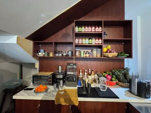 Cozinha equipada com cafeteira, liquidificador e micro-ondas. Há prateleiras com garrafas e uma cesta de frutas.