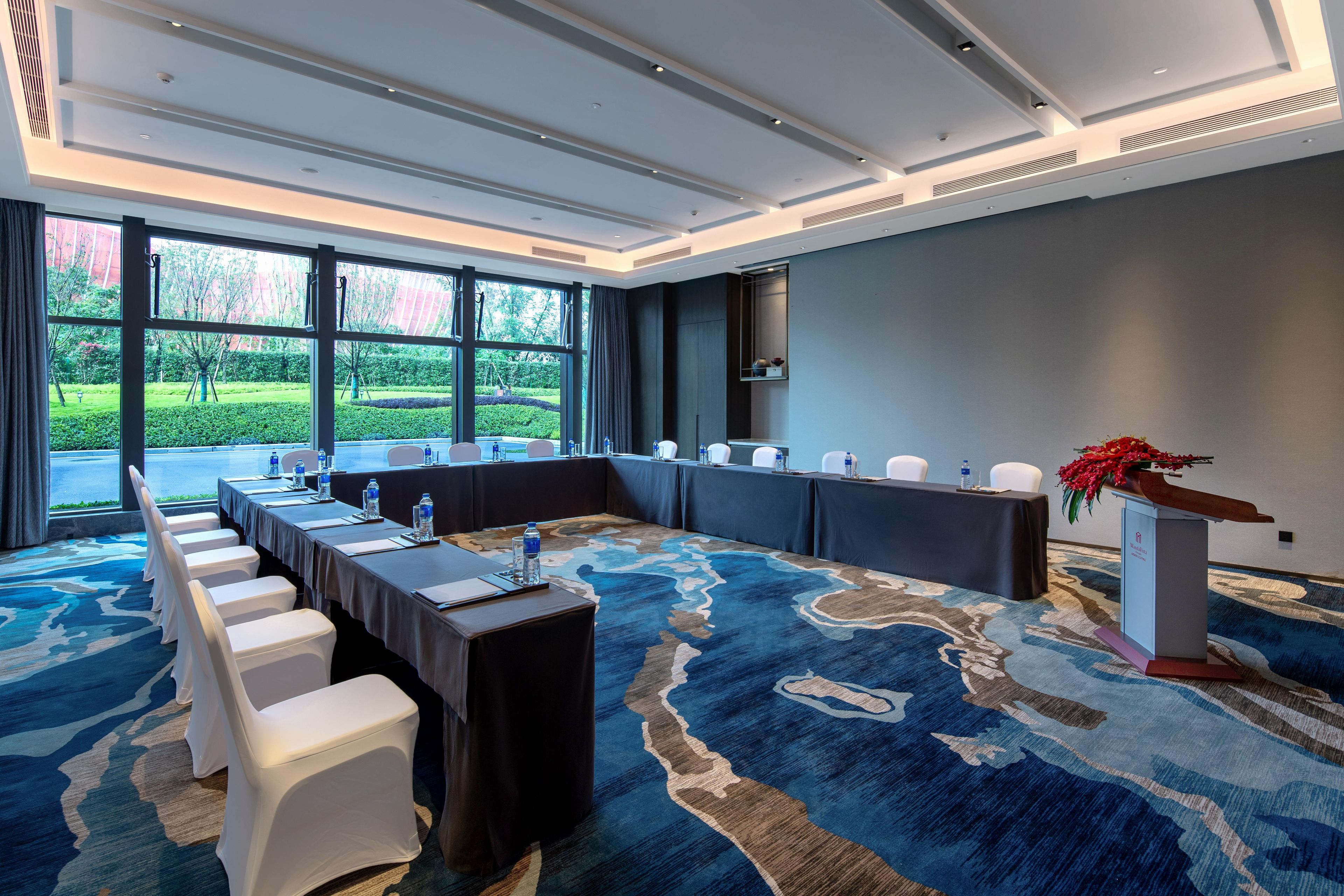 Sala de conferência com uma mesa comprida, cadeiras brancas, um pódio e uma janela grande com vista para o verde.