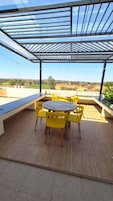Um espaço coberto ao ar livre com mesa e cadeiras, vista para o campo e céu claro.