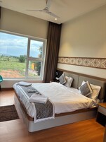 Quarto de hotel com uma cama grande, ventilador de teto, vista para o exterior e mesa de cabeceira com luminária.