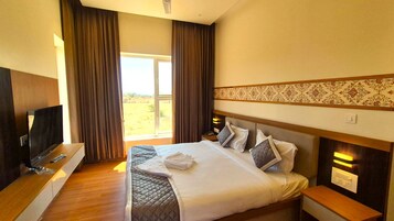 Quarto de hotel com uma cama, televisão, mesa de cabeceira de madeira e janela com cortinas.