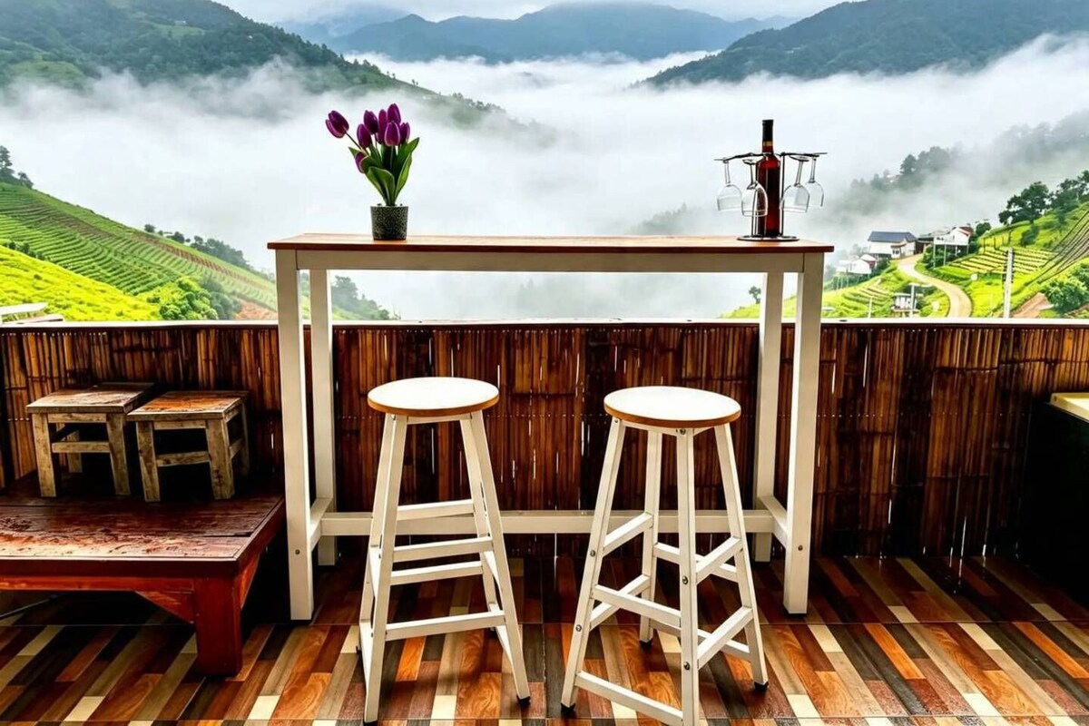 Mesa de madeira com dois banquinhos, um vaso de planta e um suporte para vinhos, com uma paisagem montanhosa ao fundo.
