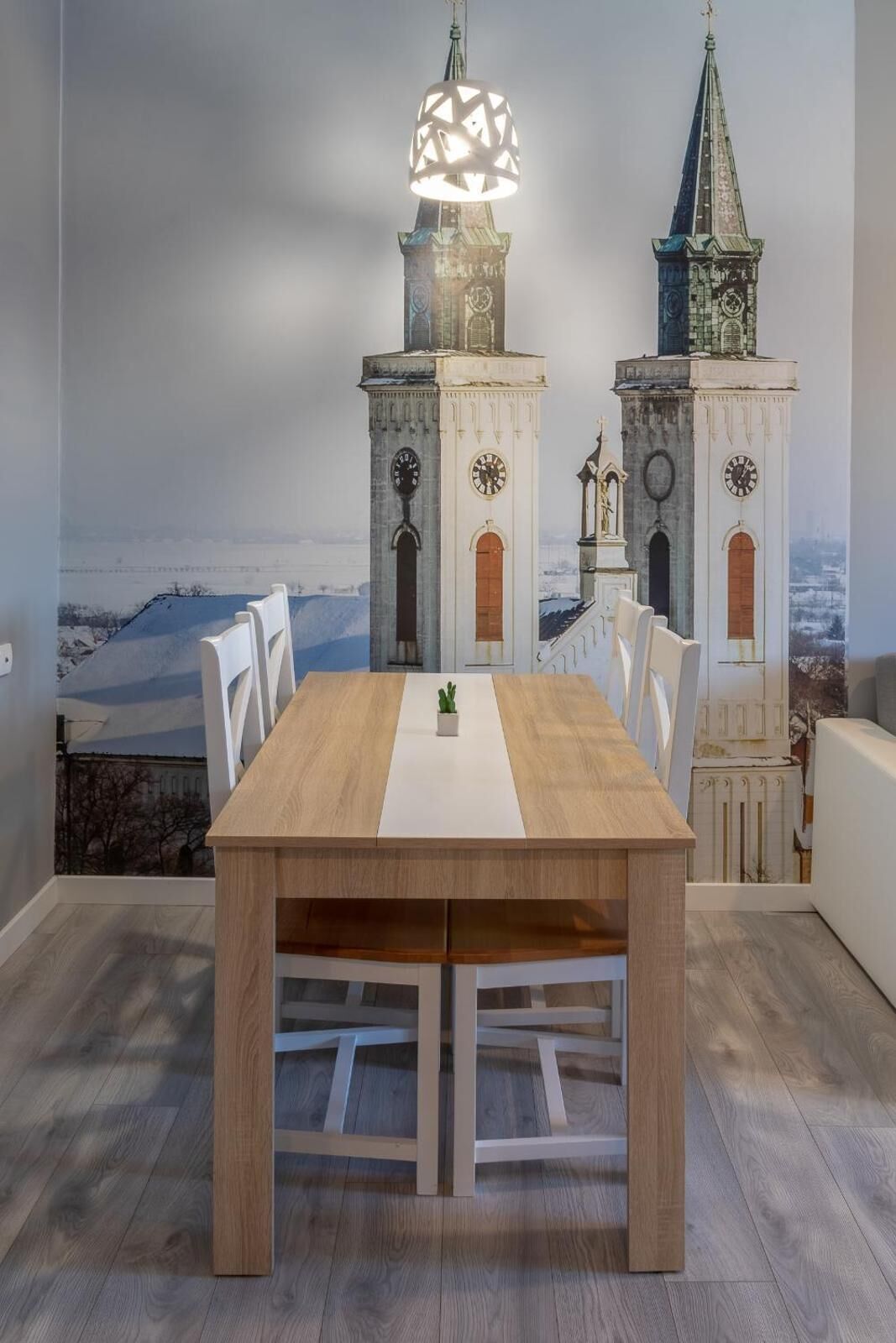 Uma sala de jantar com mesa e cadeiras de madeira, um mural na parede com duas torres e um lustre.