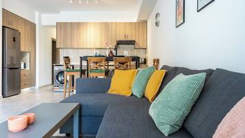 Uma sala de estar moderna com um sofá cinzento, uma mesa de centro e uma área de refeições com armários de madeira e uma cozinha ao fundo.