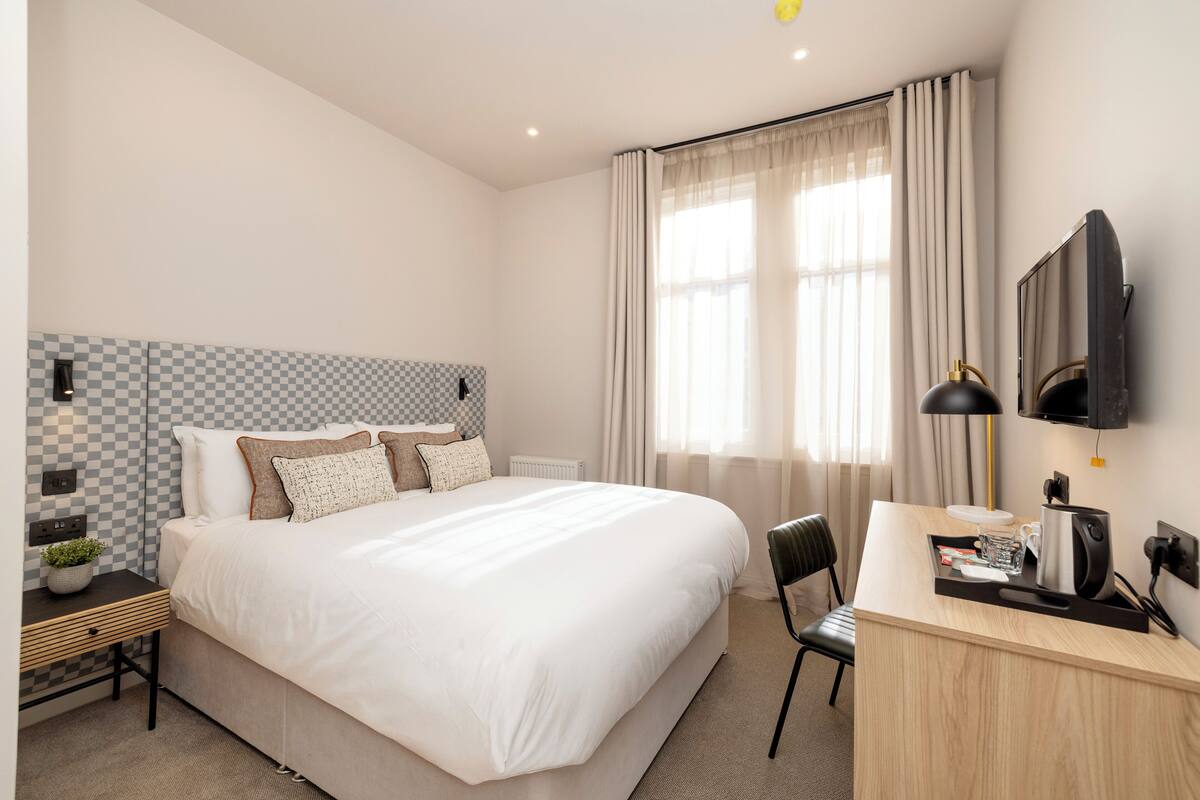 Quarto de hotel com uma cama grande, uma escrivaninha com cadeira, televisão de tela plana e uma janela com cortinas.