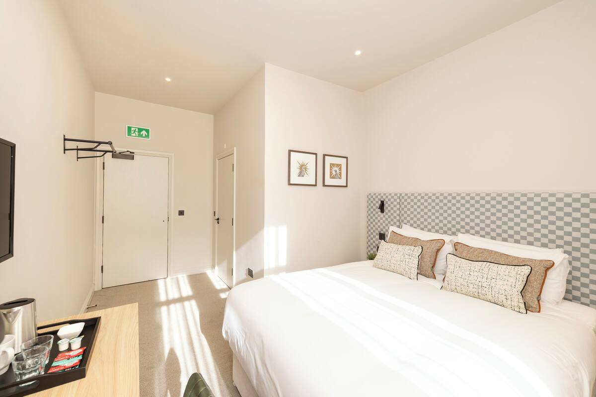 Quarto de hotel com uma cama, uma mesa de cabeceira com bandeja, uma porta com placa de saída de emergência e duas imagens emolduradas na parede.