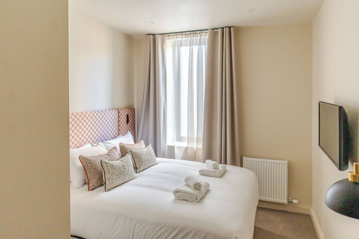 Quarto de hotel com cama, almofadas, radiador, televisão e janela com cortinas.