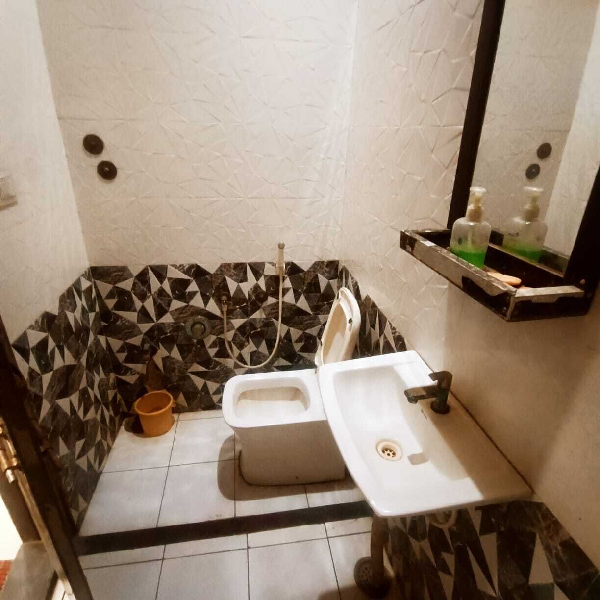 Uma casa de banho com uma pia branca, chuveiro manual, um pequeno contentor de lixo e um espelho com dispensadores de sabonete.