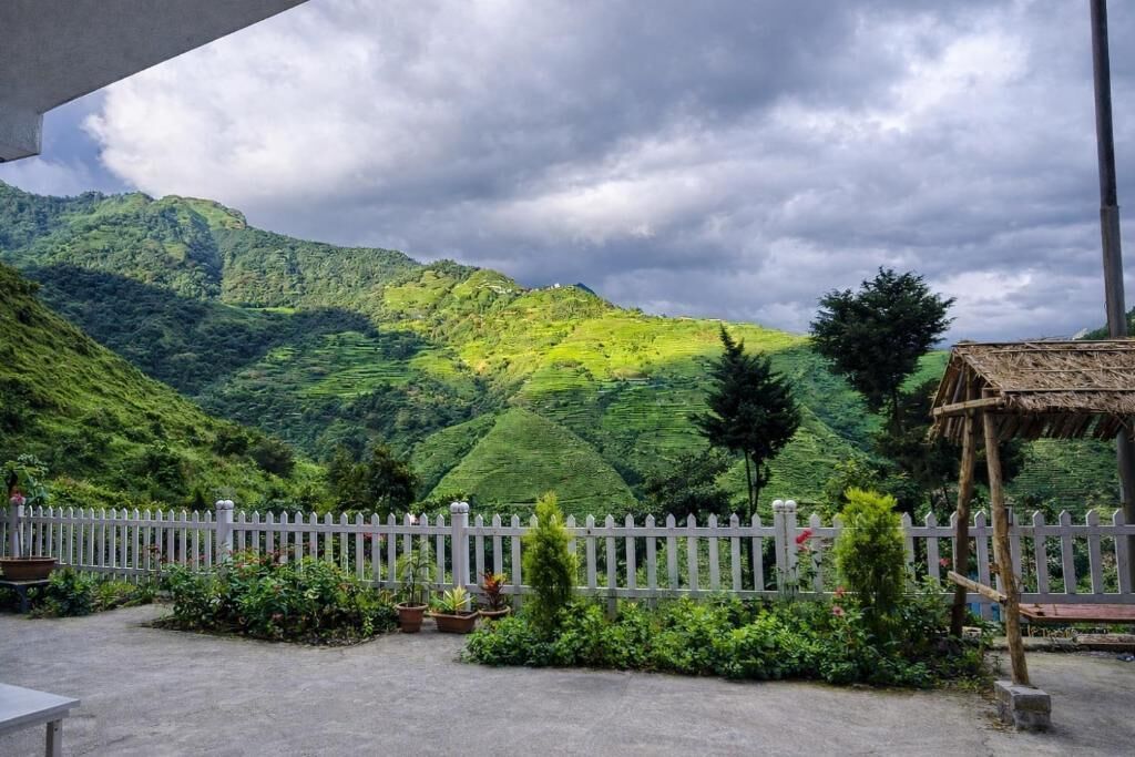 Vista de colinas em socalcos, uma cerca branca de piquete e uma construção com telhado de palha.