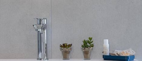Casa de banho com lavatório branco, torneira cromada, duas pequenas plantas em vasos e um prato azul com artigos de higiene pessoal.