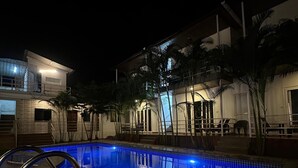 Piscina com chão de azulejos azuis e escada de metal, cercada por palmeiras e um edifício com varanda.