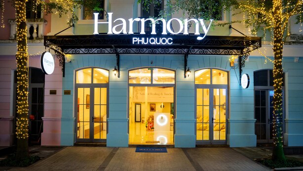 Entrada de um hotel com uma placa indicando 'Harmony PHUQUOC', apresentando um arco de boas-vindas e janelas iluminadas.