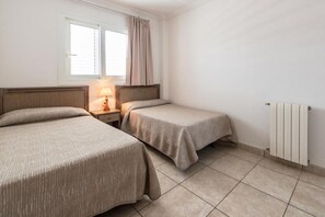 Quarto de hotel com duas camas, janela com cortinas, mesa de cabeceira com abajur e radiador.