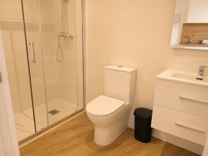 Casa de banho com cabine de duche em vidro, sanita, lavatório e um contentor de lixo.