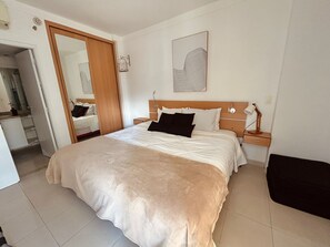 Quarto de hotel bem iluminado com uma cama grande, uma mesa de cabeceira com luminária, uma mesinha de cabeceira menor com luminária, um espelho e uma obra de arte na parede.