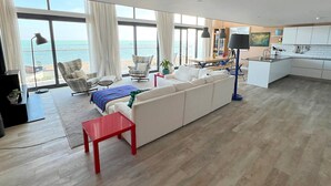 Sala moderna com um sofá branco grande, uma mesa vermelha e vista para o mar através de amplas janelas.