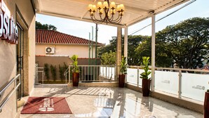 Entrada de hotel com piso de azulejos, um tapete vermelho, plantas em vasos, um candelabro e uma varanda com vista para árvores e um edifício.