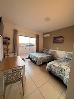 Quarto de hotel com duas camas, uma mesa de madeira, uma cadeira, uma janela com cortinas, um aparelho de ar condicionado e um quadro na parede.