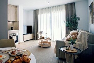 Uma sala de estar moderna com sofá, mesa de centro, mesa de jantar com itens de café da manhã, área de cozinha e uma janela grande com cortinas.