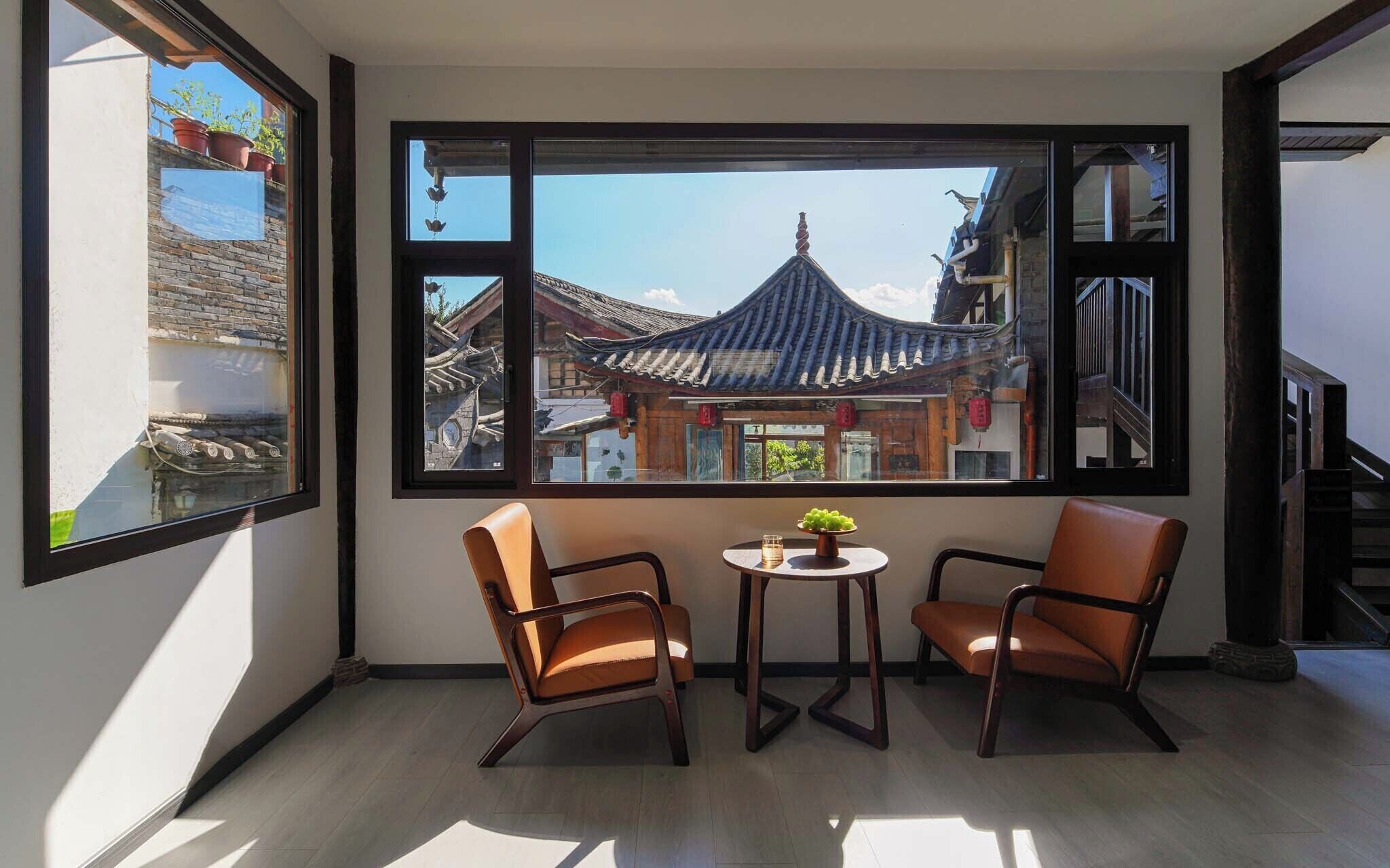 Quarto com uma grande janela, duas cadeiras, uma mesinha e vista para arquitetura tradicional.