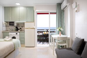 Quarto de hotel compacto com cozinha compacta, área de refeições com mesa e cadeiras, sofá e vista para o mar através de uma porta de correr.