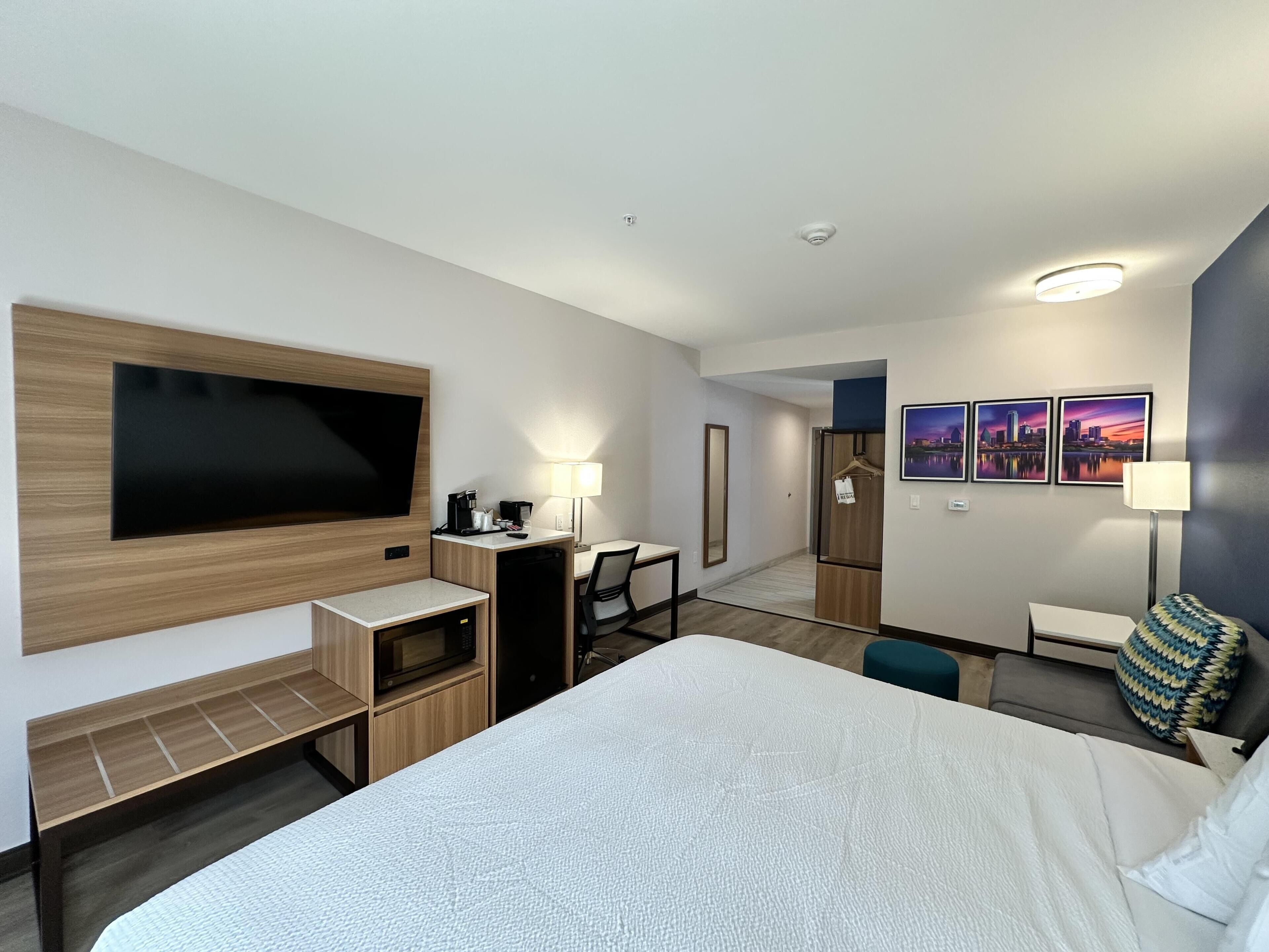 Quarto de hotel com cama, escrivaninha com computador, TV, micro-ondas, frigorífico e uma pequena área de estar.