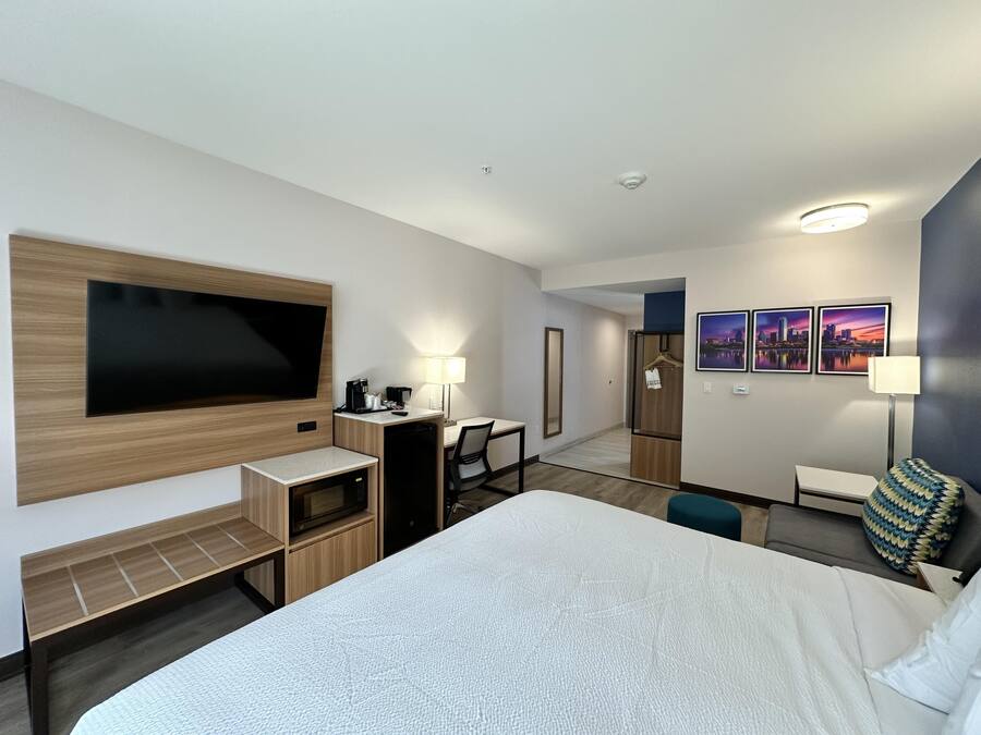 Quarto de hotel com cama, escrivaninha com computador, TV, micro-ondas, frigorífico e uma pequena área de estar.