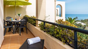 Uma varanda com mesa e cadeiras, um guarda-sol verde e vista para o mar.