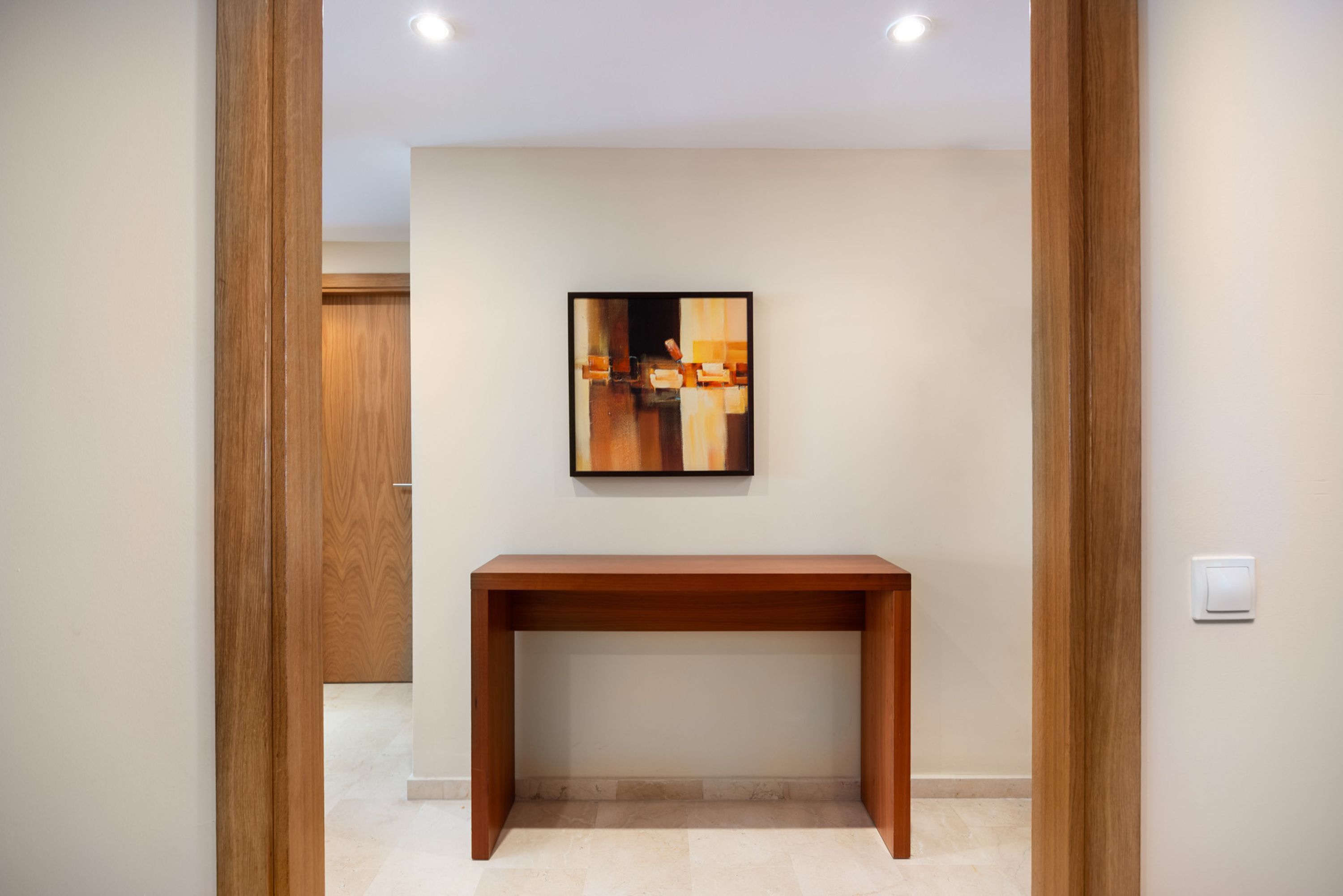 Um corredor com um aparador de madeira, um quadro abstrato emoldurado na parede e um interruptor de luz à direita.