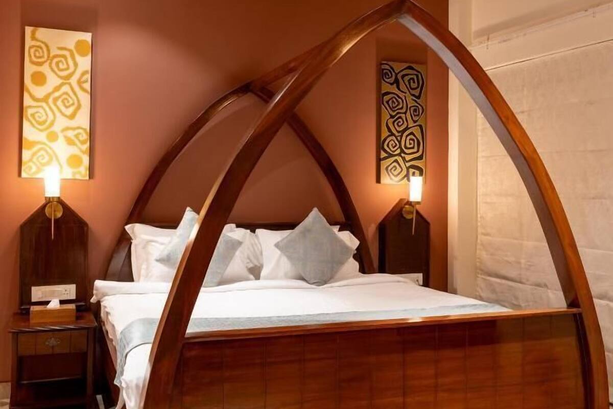 Uma cama de dossel com estrutura de madeira e roupa de cama branca, ladeada por duas mesas de cabeceira com abajures, em um quarto com painéis decorativos estampados e piso de mármore.