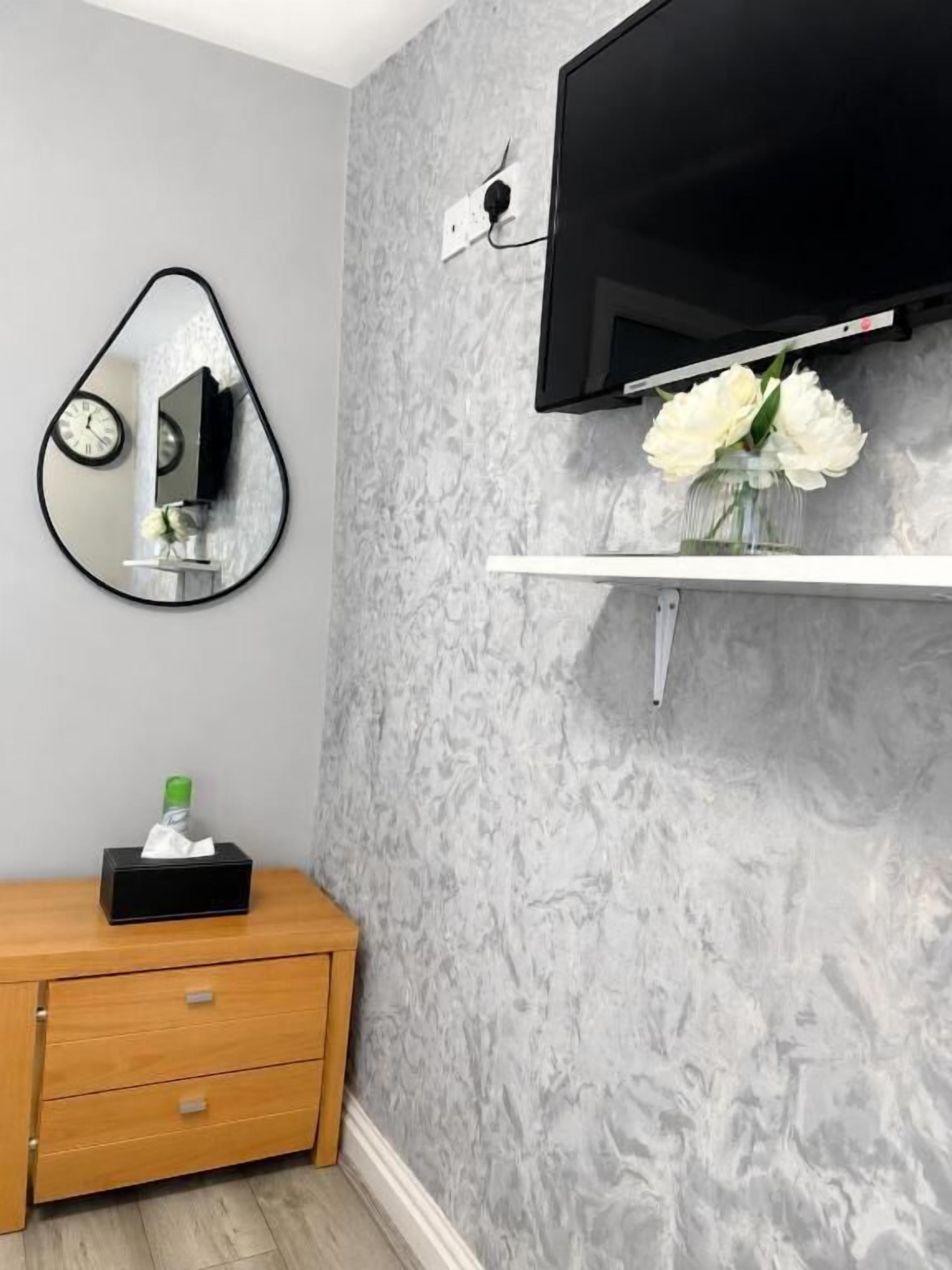Quarto com parede cinza texturizada, TV montada, mesa de cabeceira de madeira com caixa de lenços e um vaso com flores brancas numa prateleira.