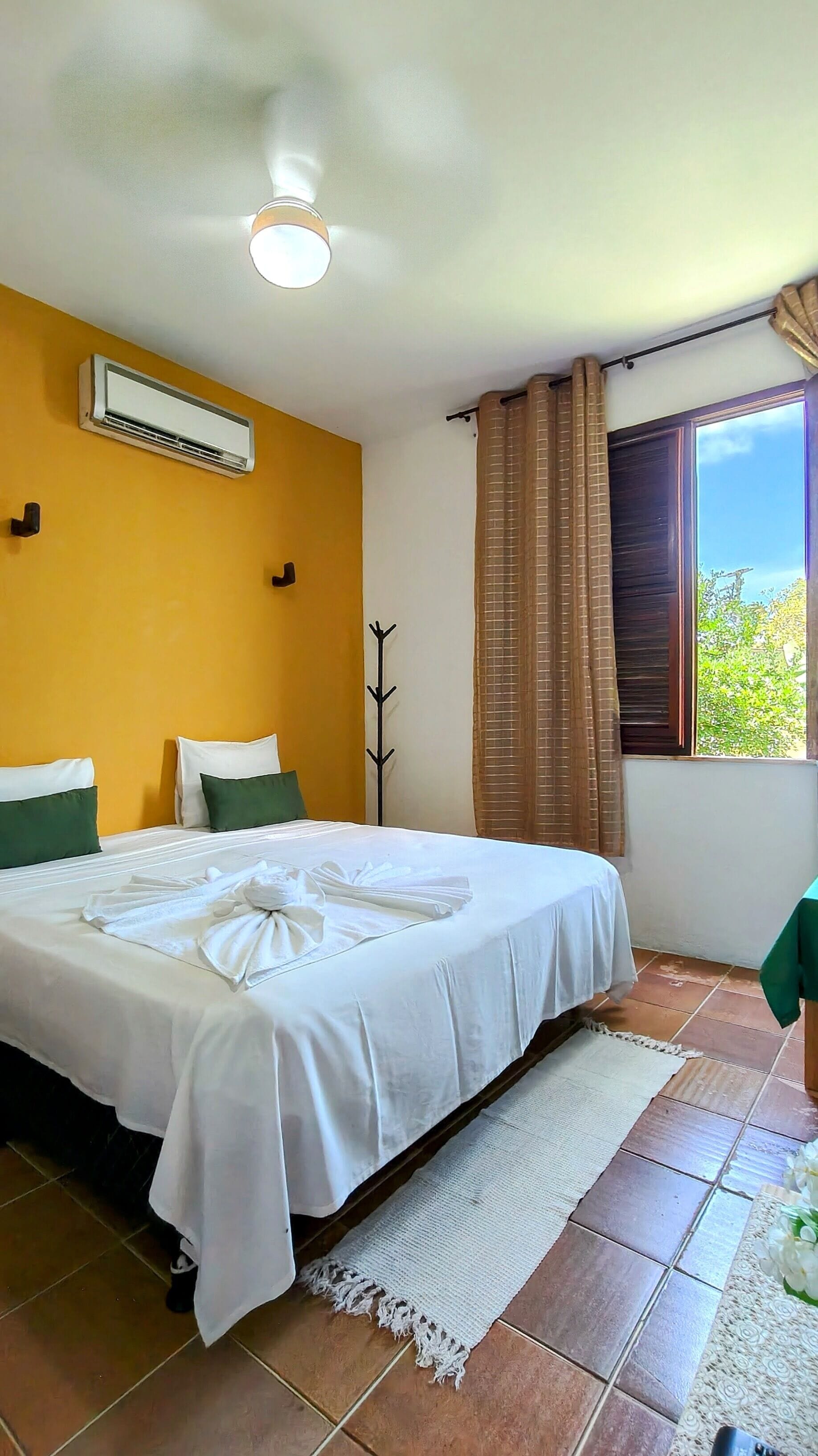 Quarto com cama, janela com cortinas, ar-condicionado fixado na parede e um cabideiro.