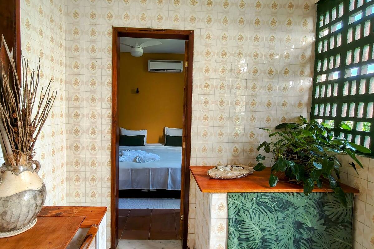 Quarto com uma cama, uma mesa de madeira, um vaso com galhos e uma planta em vaso.