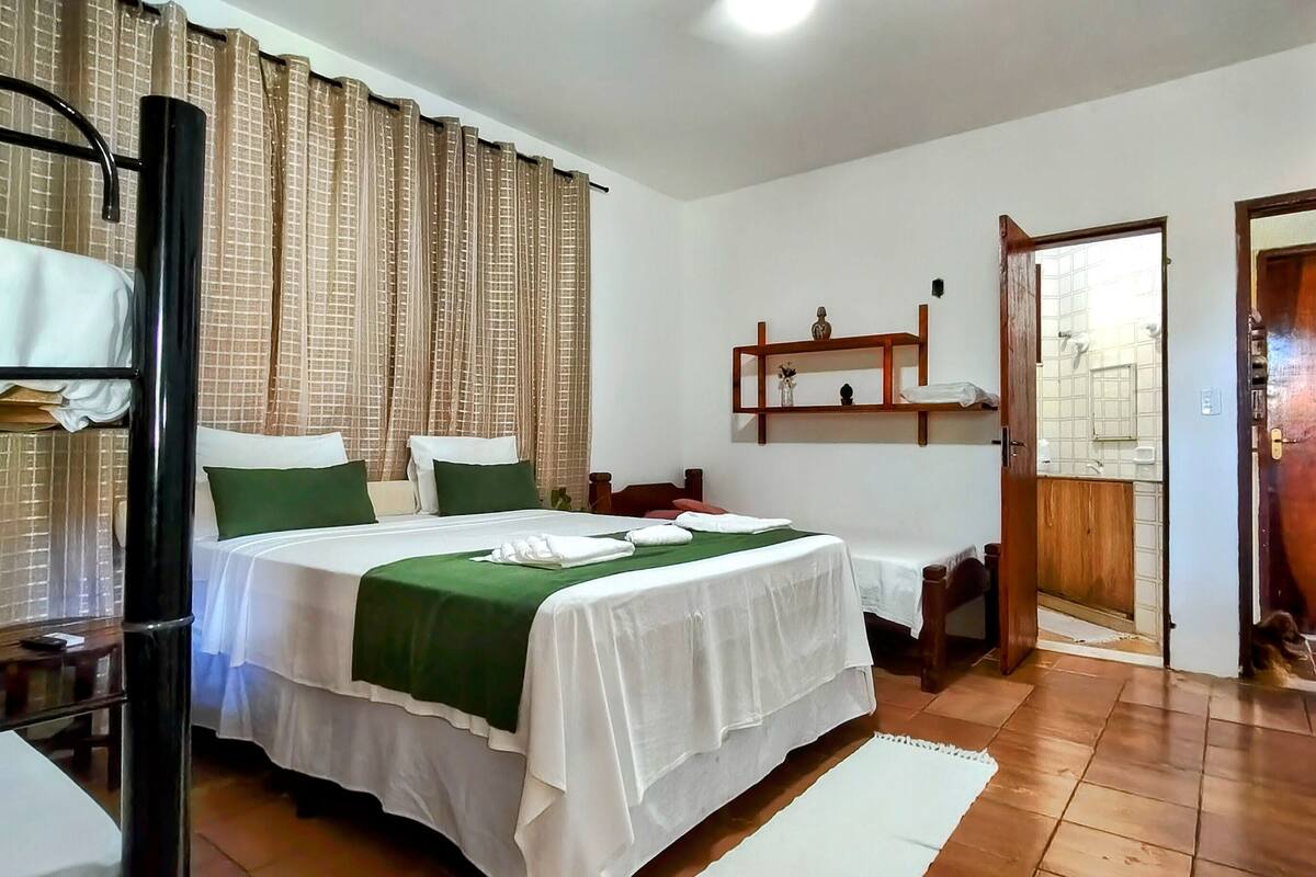Um quarto com uma cama, uma mesinha de cabeceira, uma estante com objetos decorativos, uma porta e uma janela com cortinas.