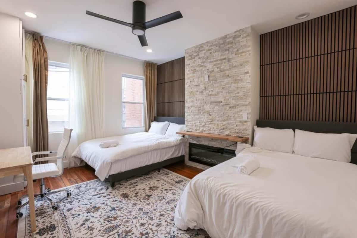 Quarto com duas camas, uma escrivaninha, uma cadeira, um ventilador de teto e uma parede de tijolos.