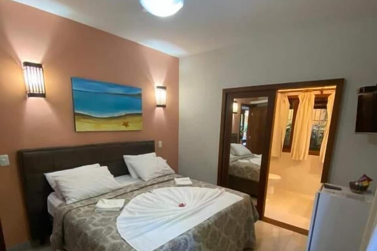Quarto de hotel com cama, ventoinha no teto, um quadro na parede e porta que dá para outro cômodo.