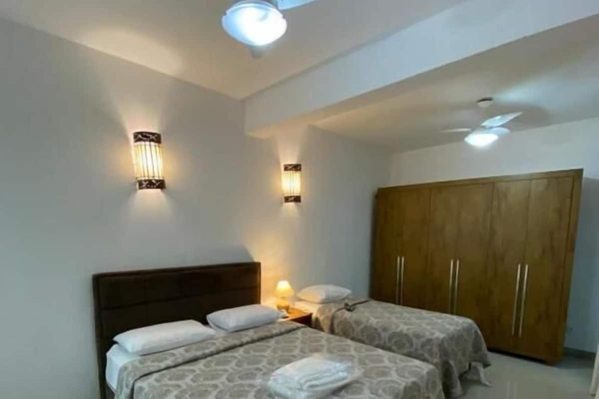 Quarto de hotel com duas camas, guarda-roupa, ventoinha no teto e luzes fixadas na parede.