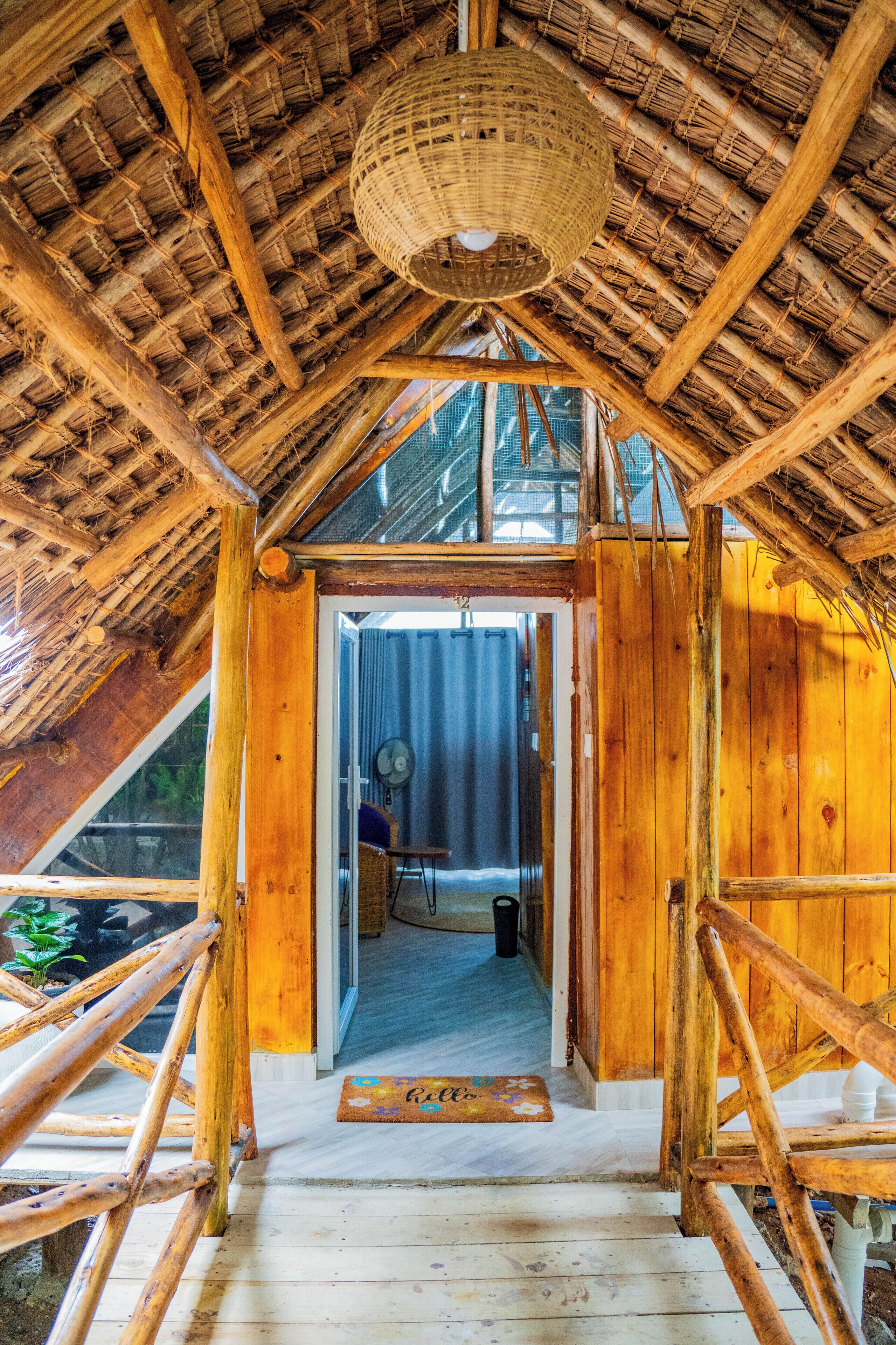 Quarto de sótão com telhado de colmo, vigas de madeira, luminária de cesta trançada e vista pela janela.