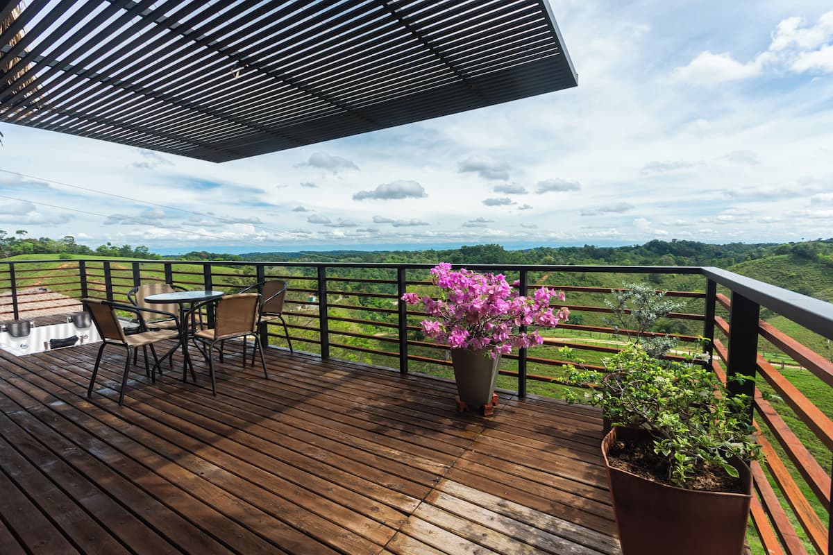 Um deck de madeira com mesa e cadeiras, um vaso com flores cor-de-rosa e vista para campos verdes.