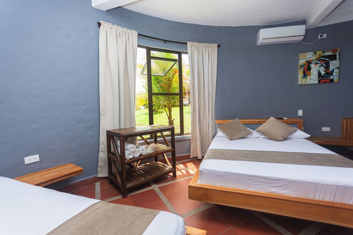 Quarto com duas camas, um banco de madeira com toalhas, uma janela com cortinas e um quadro na parede.