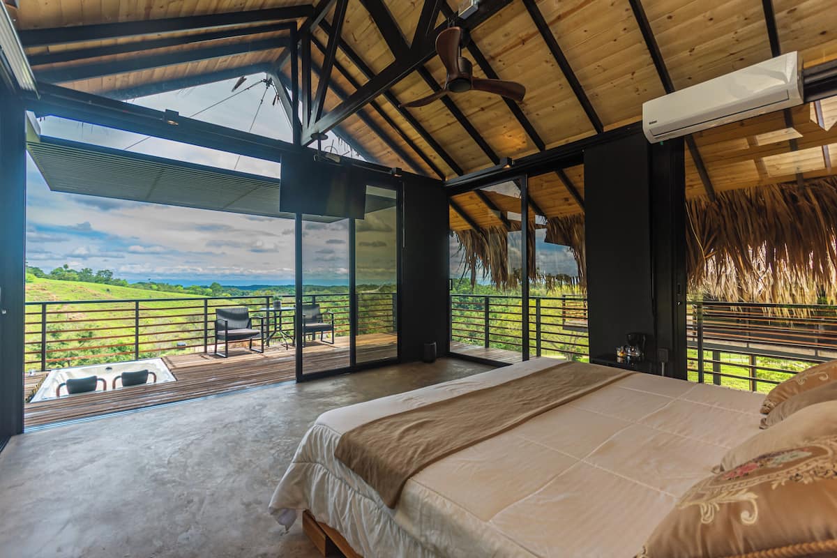 Quarto com uma cama grande, vista para o campo, varanda com mesa e cadeiras e um ventilador de teto.