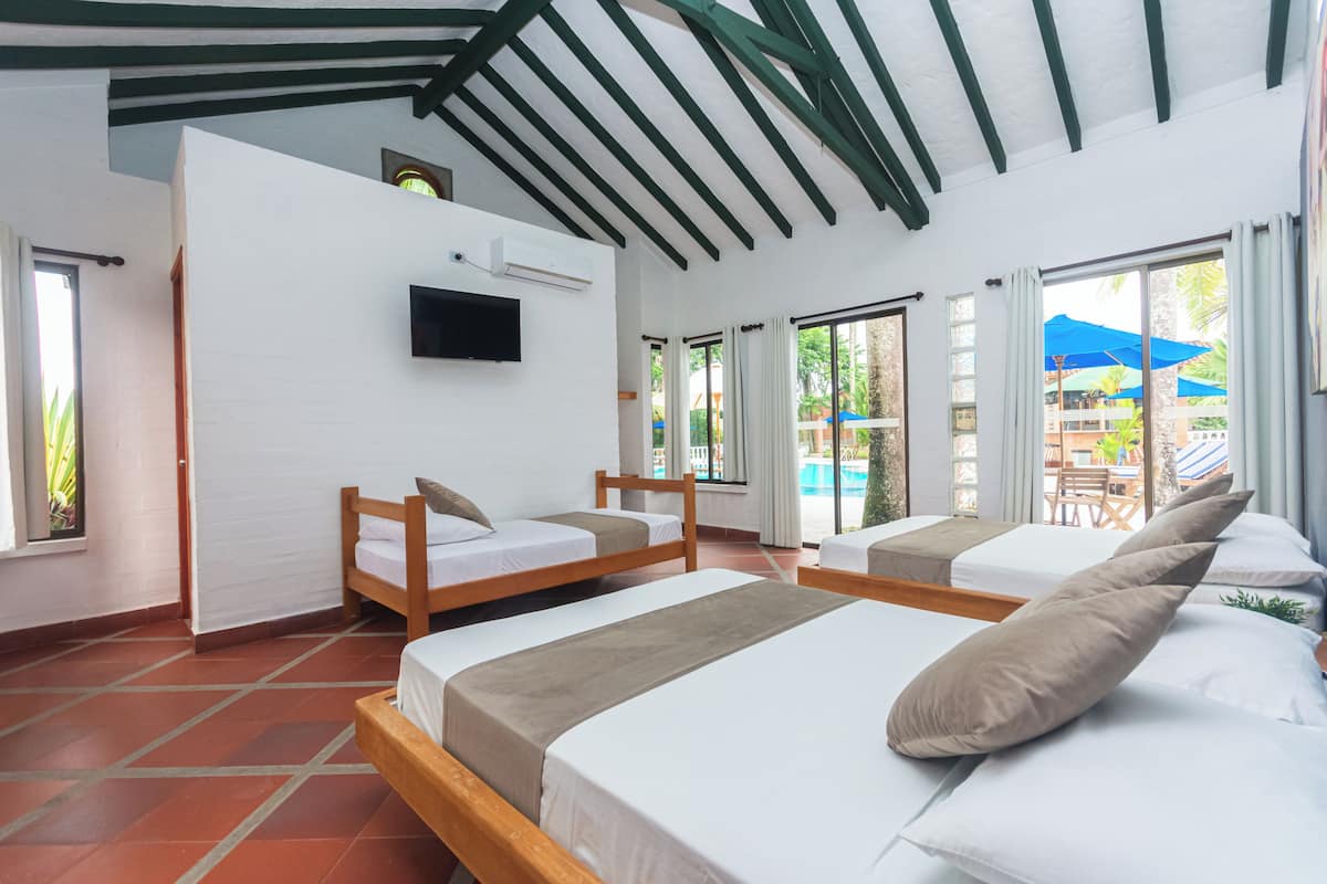 Quarto de hotel com três camas de madeira, uma televisão de tela plana e janelas amplas com cortinas.