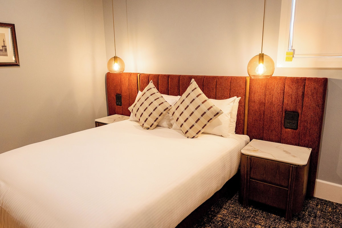 Uma cama bem arrumada com cabeceira de madeira, duas mesas de cabeceira e duas luminárias pendentes.