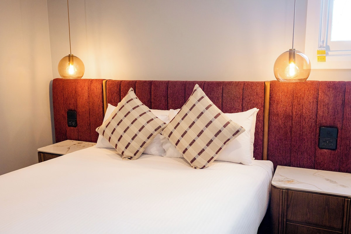 Cama com roupa de cama branca, duas mesas de cabeceira, painel de cabeceira de madeira e duas luminárias pendentes.