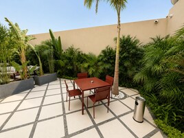 Um pátio externo com mesa e cadeiras, cercado por vegetação exuberante e piso de azulejos.